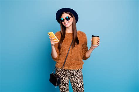 긍정적 인 쾌활한 소녀 냉각 사용 스마트 폰 팔로우 공유 Repost 읽기 블로거 보유 머그컵 카페인 음료 착용 갈색 바지 복고풍 빈티지 모자 고립 된 파란색 색상 배경