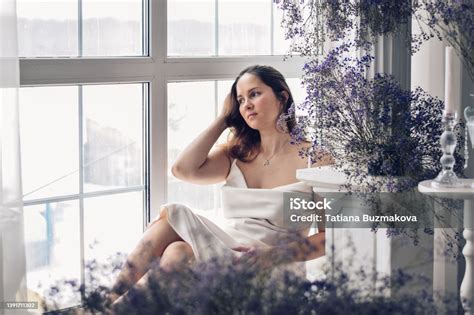 가벼운 실크 드레스를 입은 아름다운 젊은 장발 여성이 2022 년의 석고 필라 트렌디 한 색상으로 장식 된 방의 탁 트인 창문을 바라 봅니다 2022년에 대한 스톡 사진 및