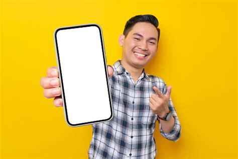 하얀 체크무늬 셔츠를 입고 웃고 있는 잘생긴 젊은 아시아 남성이 빈 화면으로 휴대폰을 가리키며 노란색 배경에 격리된 새로운 모바일 앱을 광고하고 있다 사람들의 라이프스타일