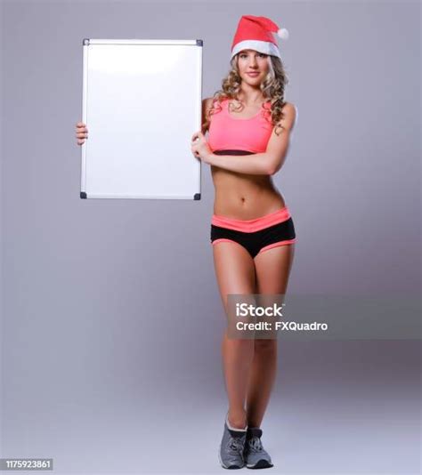 스튜디오에서 빈 보드와 함께 포즈를 취하는 스포츠웨어와 산타의 모자를 입고 아름다운 피트니스 모델 회색 배경 위에 격리되었습니다 2019년에 대한 스톡 사진 및 기타 이미지