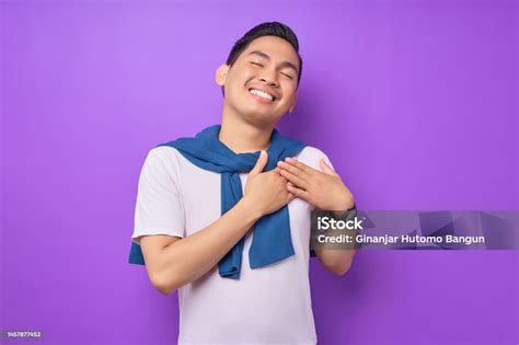 흰색 티셔츠를 입고 보라색 배경 위에 고립된 가슴에 손바닥을 교차시킨 채 눈을 감고 있는 화난 젊은 아시아 남자를 즐기십시오