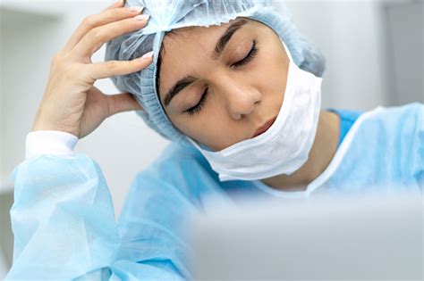 얼굴 마스크와 수술 용 바지를 입은 피곤한 여성 의사가 눈을 감고 있는 의료 사무실의 노트북 화면 앞에 앉아 있습니다 의료 및 의료 개념 25 29세에 대한 스톡 사진 및