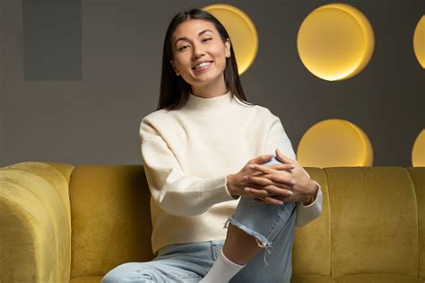 행복한 아시아 여성은 카메라를 바라보며 대화를 나누고 화상 콜 컨퍼런스 노란 소파에 앉아있는 집에서 온라인 인터뷰를 개최합니다