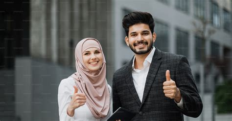 히잡을 쓴 웃는 여자와 사무실 건물 근처 야외에 서 있는 동안 엄지손가락을 치켜든 무슬림 잘생긴 남자 두 비즈니스 파트너의