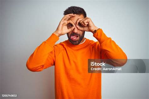 고립 된 흰색 배경 위에 오렌지 스웨터를 입은 젊은 인도 남자는 혀를 내밀고 손가락을 통해 눈을 내밀고 있는 쌍안경처럼 괜찮은 제스처를하고 있습니다 미친 표정 교도소에 대한