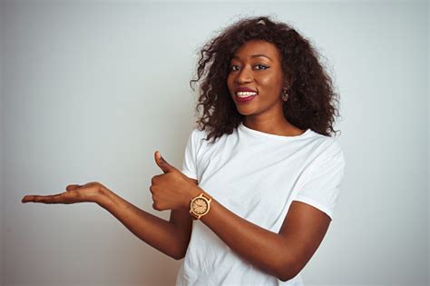 고립 된 흰색 배경 위에 서있는 티셔츠를 입은 젊은 아프리카 계 미국인 여성은 손바닥 손을 표시하고 엄지 손가락으로 확인 제스처를하고 행복하고 쾌활한 미소를 지었습니다