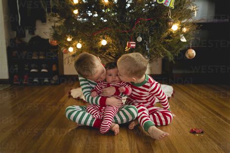 Brothers Kissing Sister While Sitting Against Christmas Tree At Home