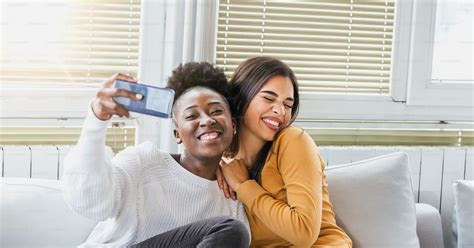 웃고 있는 여자 친구들은 스마트폰으로 셀카 사진을 찍는 동안 소파에서 서로 껴안고 있다 사랑스러운 백인과 아프리카계 미국인