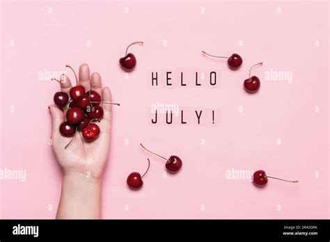 Hello July Text And Female Hand Holds Ripe Cherries On Pink Background
