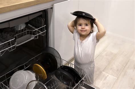 식기 세척기를 내리는 데 도움이되는 작은 귀여운 유아 소녀 집에서 흰색 부엌에서 재미있는 작은 엄마의 도우미 건강한 아이는 집안일을하고 있습니다 부엌에서 즐기는 아이 식기