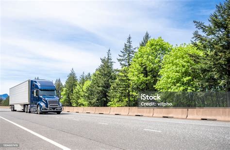 다크 블루 스타일리시 빅 리그 세미 트럭 트랙터와 그릴 가드가 고정된 화물을 운반하는 리퍼 세미 트레일러에서 숲이 있는 넓은 고속도로 도로에서 달리는 세미 트럭에 대한 스톡