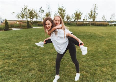 행복한 엄마와 어린 소녀가 공원에서 함께 놀고 즐거운 시간을 보내고 있습니다 소녀는 흰색 티셔츠를 입은 어머니의 등을 타고 프리미엄 사진