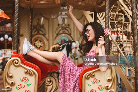 선글라스와 드레스에 어두운 곱슬 머리를 가진 쾌활한 아가씨는 손에 롤리 팝 사탕을 들고 놀이 공원에서 시간을 보내는 동안 회전 목마를 타고 20 24세에 대한 스톡 사진 및