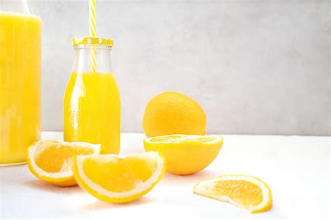 뚜껑이 달린 유리 병과 오렌지 주스가 들어간 칵테일 튜브가 회색 콘크리트 벽에 흰색 테이블 위에 서 있습니다 육즙이 많은 오렌지 조각이 근처에 있습니다 해독 다이어트 음료