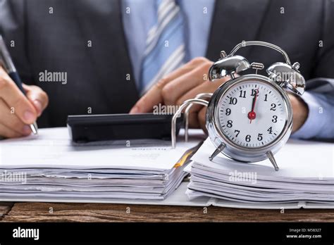 Midsection Of Businessman Calculating Invoice With Alarm Clock On Documents At Table Stock Photo