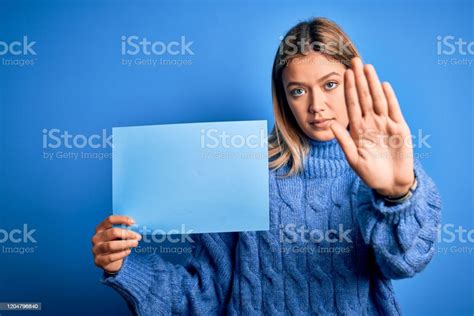 진지하고 자신감있는 표정 방어 제스처로 정지 표지판을하고 열린 손으로 고립 된 파란색 배경 위에 서있는 배너를 들고있는 젊은 아름다운 여성 갈색 머리에 대한 스톡 사진 및