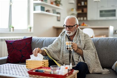 독감에 걸리고 낮에는 거실 소파에서 뜨거운 차를 마시는 아픈 노인 독감에 걸린 감기에 지친 노인은 따뜻한 담요에 싸여 따뜻한 차 한 잔을 들고 거실에서 집에서 마신다 65