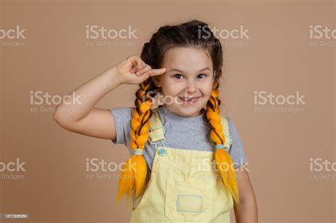노란 카네칼론 피그테일을 들고 흥분한 빛나는 어린 소녀 노란 점프수트와 베이지 색 배경에 회색 티셔츠를 입고 사원으로 손가락을 들고 실종된 이빨로 미소 짓고 있다 땋은