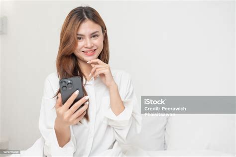 하얀 잠옷을 입은 여자가 주말 아침에 일어나 휴식을 취하고 노트북 휴대폰을 가지고 놀며 하얀 침실 안의 유리에서 빵을 먹고 차를 마신다 아침 휴가 개념입니다 갈색 머리에