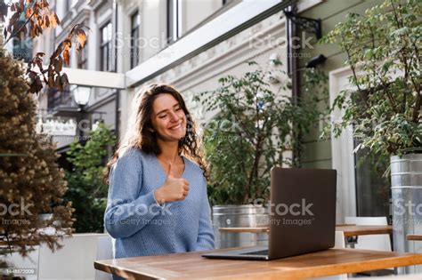 노트북을 들고 엄지손가락을 치켜들고 테라스의 카페에서 야외 비디오 연결을 통해 동료들과 채팅하는 행복한 소녀 쾌활한 젊은 여성은 친구 및 가족과 함께 온라인에서 즐거운