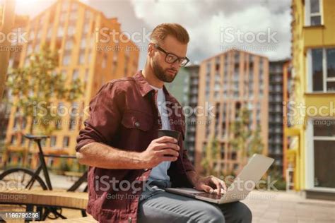 커피가 필요해요 야외 벤치에 앉아있는 동안 캐주얼 옷과 안경에 수염을 가진 잘 생긴 남자 노트북에서 작업하고 커피 한 잔을 들고