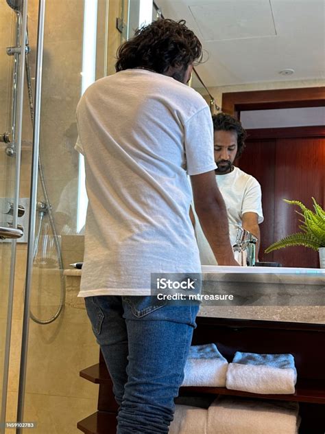 인도 남자의 거울에 비친 반사의 클로즈업 이미지는 흐르는 물에서 손을 씻는 고급 욕실의 싱크대에 서 있었고 대리석 욕실 카운터의 금속 냄비에 인공 양치류 식물 흐르는 수도