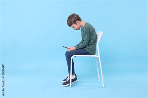 Boy With Incorrect Posture And Phone Sitting On Chair Against Light