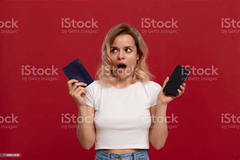 빨간 배경에 흰색 티셔츠에 곱슬 금발 머리를 가진 여자의 초상화입니다 깜짝 놀란 모델은 카메라를 보고 휴대 전화로 여권을 들고