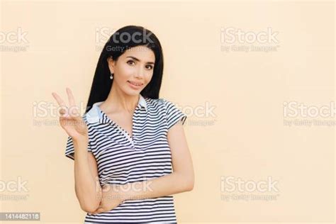 매력적인 갈색 머리 여성의 스튜디오 샷 기쁘게 표현 제스처 실내 및 평화 기호를 보여줍니다 메이크업과 깨끗한 순수한 피부를 가지고 캐주얼 옷을 입고 스튜디오 벽에 고립 25