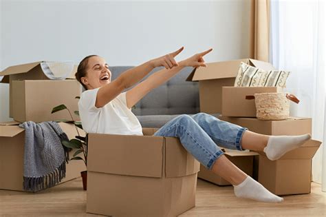 하얀 티셔츠와 청바지를 입고 골판지 상자에 앉아 상자 검지 손가락으로 가리키며 즐겁고 놀란 무언가를 보여주는 흥분한 여성의 수평 촬영 30 34세에 대한 스톡 사진 및 기타