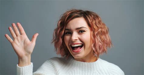 초상화 행복한 젊은 여자는 놀람에서 군중 속의 사람들을보고 친구들의 익숙한 얼굴을보고 그들에게 미소를 짓고 손을 흔들어 인사합니다 감정 모델 회색 배경입니다 공간을