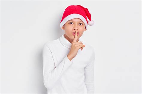 흰색 티셔츠를 입고 산타클로스 모자를 쓴 진지한 젊은 아시아 아이가 흰색 배경에 고립된 침묵을 유지하도록 요청하는 쉿 제스처를 보여줍니다 메리 크리스마스와 새해 복 많이