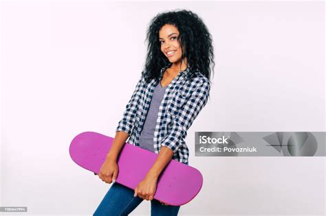 젊고 스포티합니다 캐주얼 한 옷을 입은 성인 아프리카 계 미국인 소녀가 보라색 스케이트 보드를 손에 들고 저항 할 수없는 미소로 카메라를 바라보고 있습니다 가냘픈에 대한