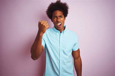 고립 된 분홍색 배경 에 서 파란색 셔츠를 입고 아프리카 머리를 가진 젊은 미국 남자는 분노와 함께 외치는 동안 좌절과 분노를 제기 주먹 분노와 공격적인 개념 감정에 대한