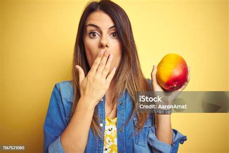 노란 배경 커버 입에 신선한 건강 망고를 먹는 젊은 아름 다운 여자 실수에 대 한 수치감 두려움의 표정 침묵에 무서워 비밀 개념 가리기에 대한 스톡 사진 및 기타 이미지