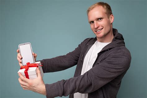 잘 생긴 행복 한 젊은 금발 남자 고립 된 파란색 배경 벽에 캐주얼 세련 된 옷을 입고 흰색 선물 상자를 들고 빨간 리본과 휴대 전화 와 빈 화면 디스플레이 카메라를 보고