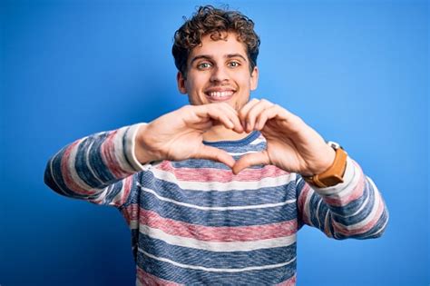 젊은 금발 잘생긴 남자 곱슬 머리 를 입고 줄무늬 스웨터 위에 파란색 배경 미소 사랑 보여주는 심장 기호와 손으로 모양 로맨틱 한 개념 개념에 대한 스톡 사진 및 기타