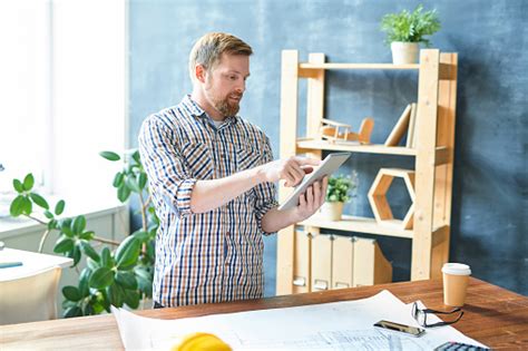 기술 도면 및 워크샵에서 태블릿을 사용 하 여 책상에서 서 있는 체크 무늬 셔츠에 명랑 자신감 잘생긴 젊은 수염 엔지니어 건설 산업에 대한 스톡 사진 및 기타 이미지