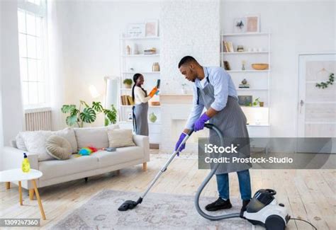 그의 아내는 거실 여유 공간에서 선반을 청소하는 동안 젊은 흑인 남자 진공 청소기 바닥의 전체 길이 2명에 대한 스톡 사진 및