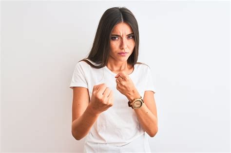 고립 된 흰색 배경 위에 서있는 캐주얼 한 티셔츠를 입은 젊은 아름다운 여성은 주먹 방어 제스처 화가 나고 화가 난 얼굴 문제를 두려워하여 싸울 준비가되어 있습니다 T