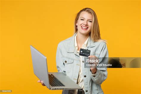 노트북 Pc 컴퓨터 스튜디오에서 노란색 오렌지 벽 배경에 고립 된 신용 은행 카드를 들고 데님 캐주얼 옷에 웃는 젊은 여자의 초상화 사람들 라이프 스타일 개념입니다 복사
