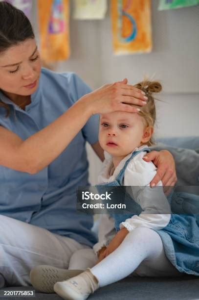 교실 소파에 앉아 있는 어린 소녀의 이마에서 열이 나는지 걱정스러운 간병인이 몸이 좋지 않아 보살핌과 위로가 필요한 것처럼 보입니다 2명에 대한 스톡 사진 및 기타 이미지