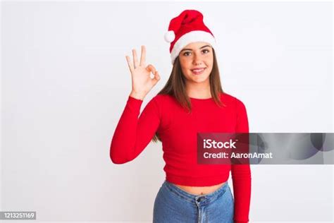 고립 된 흰색 배경 위에 서있는 크리스마스 산타 모자를 쓰고있는 어린 아름다운 소녀는 손과 손가락으로 확인 기호를하고 긍정적 인 미소를 짓고 있습니다 성공적인 식입니다 12