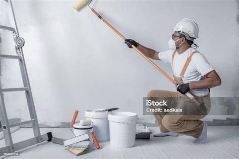 한 남자가 롤러로 하얀 벽을 그립니다 내부의 수리 빈 방에 벽을 그리는 젊은 남성 데코레이터 개념 빌더 또는 빈 방에 페인트 롤러와 헬멧에 화가 Diy에 대한 스톡 사진 및