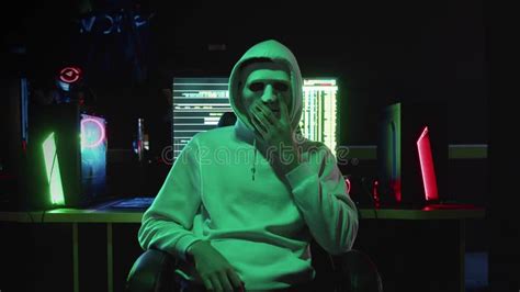 Portrait Of A Mysterious Hacker In A Hood Sitting At A Table And Looking At The Camera After