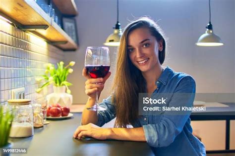 아름 다운 귀여운 젊은 캐주얼 미소 행복한 술 마시는 여자의 초상화 레드 와인 한 잔을 들고 집에 있는 로프트 스타일의 부엌에 테이블에 앉아 있다 와인에 대한 스톡 사진 및