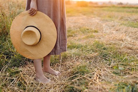 모자를 손에 든 여자는 일몰 빛으로 짚밭에서 맨발로 걷고 자른 전망 시골의 느린 삶 대기의 고요한 순간 소박한 린넨 드레스를 입은 젊은 여성이 여름 시골에서 휴식을 취합니다