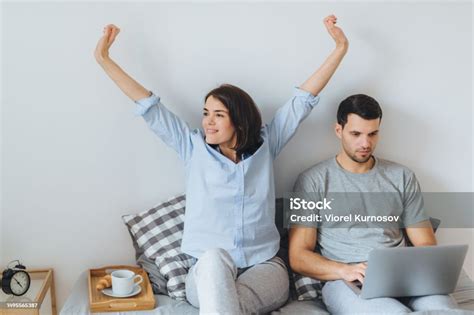 젊은 부부 침대에서 아침 지출 아름 다운 갈색 머리 여성 뻗어와 심각한 표정으로 그녀의 남편 사용 노트북 컴퓨터 원격 작업 부담