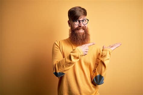 노란색 고립 된 배경위에 안경을 쓰고 수염을 기르는 잘 생긴 아일랜드 빨간 머리 남자는 손으로 손가락으로 가리키는 동안 카메라에 놀랍고 미소를 지었습니다 노랑에 대한 스톡