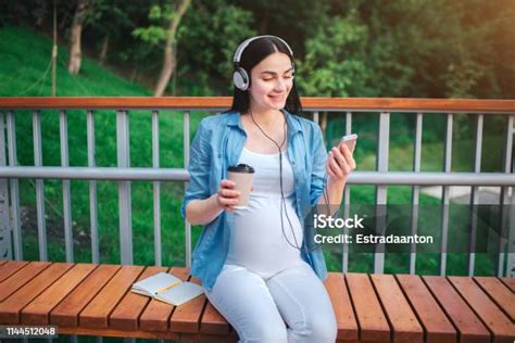 배경의 도시에서 행복 한 검은 머리와 자랑 임신한 여자의 초상화 그녀는 도시 벤치에 앉아 있다 임산부와 함께 공원에서 음악을 듣는 엄마 거리에 대한 스톡 사진 및 기타